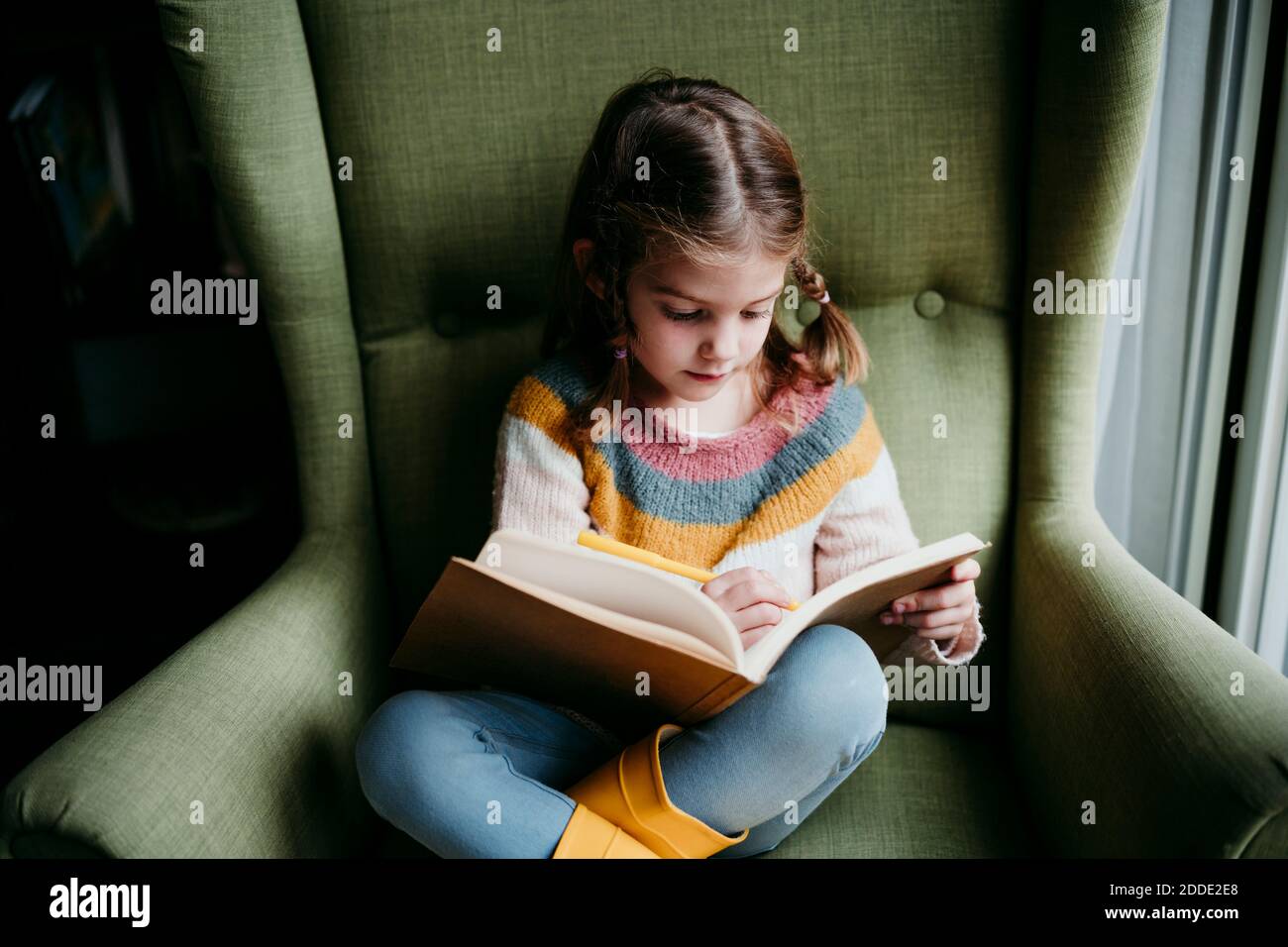 Cute girl writing in book while sitting on chair at home Stock Photo ...