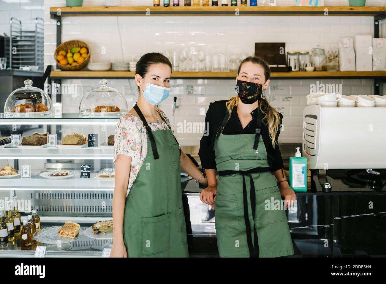 Partners wearing face mask and apron standing by counter at coffee shop ...