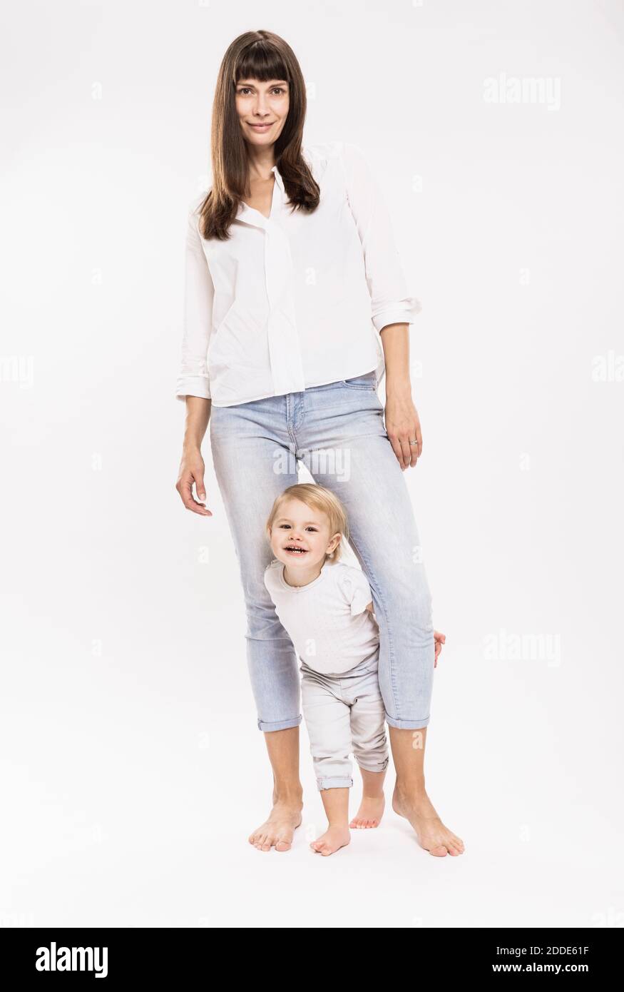 Daughter standing in between of mother's leg against white background ...