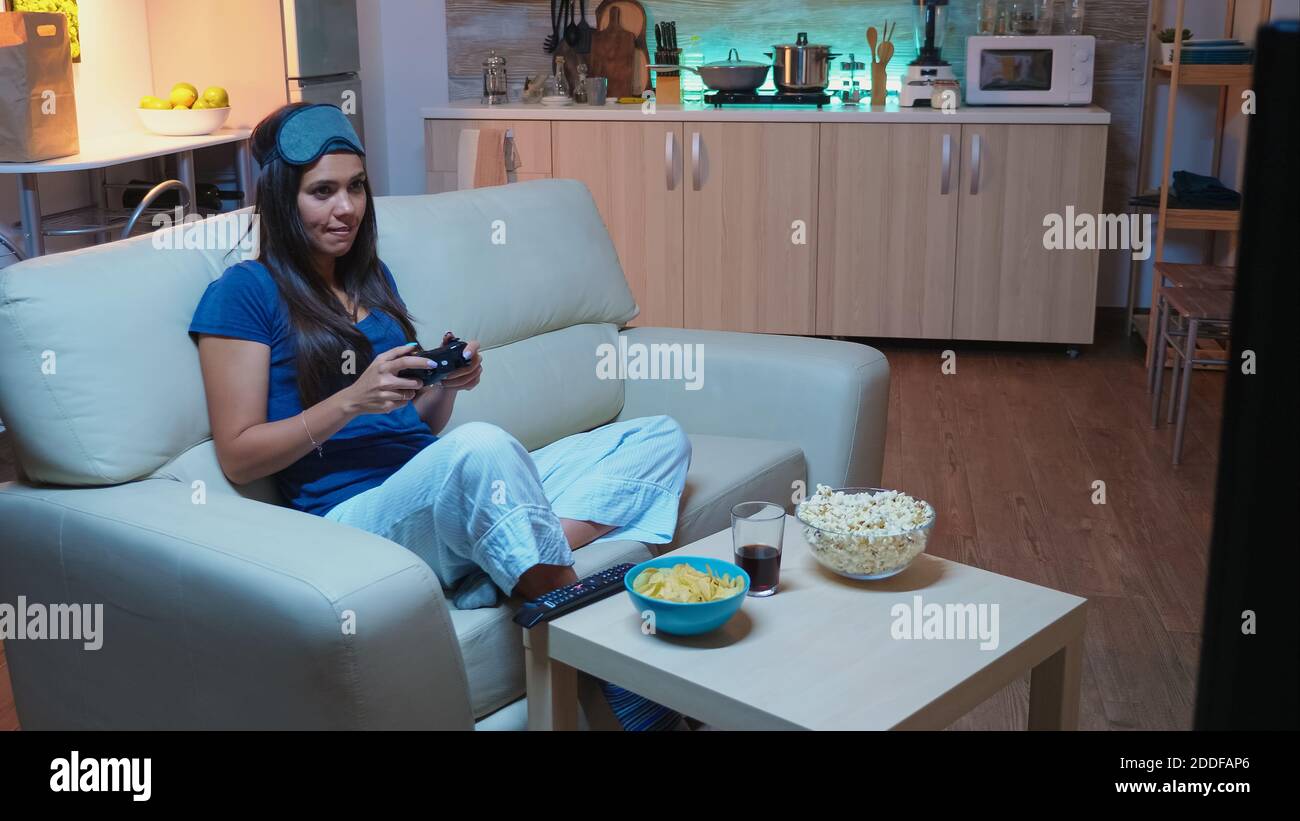 Woman sitting on sofa playing video game late at night wering eye mask ...
