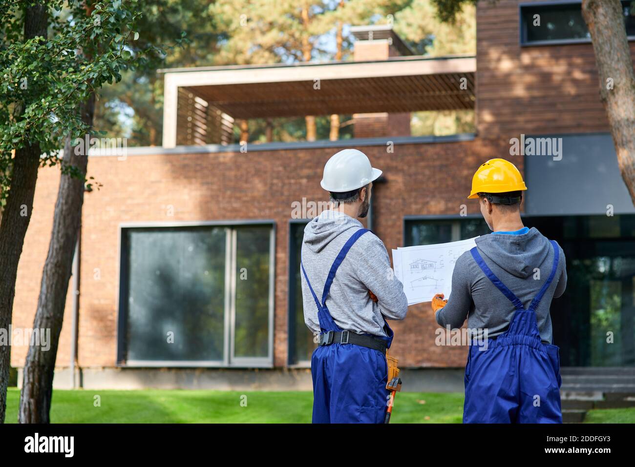 Rear view of two builders standing outdoors with an open blueprint ...