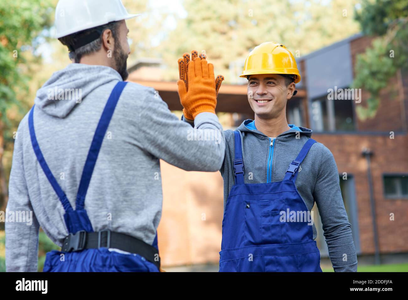 Two positive young male engineers in hard hats smiling at each other ...
