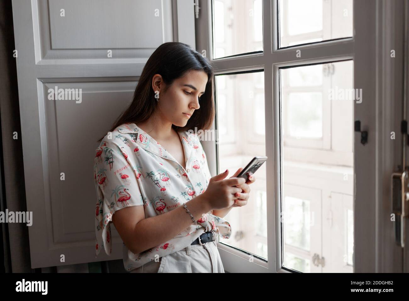 Side view of calm female browsing on smartphone standing near window at ...