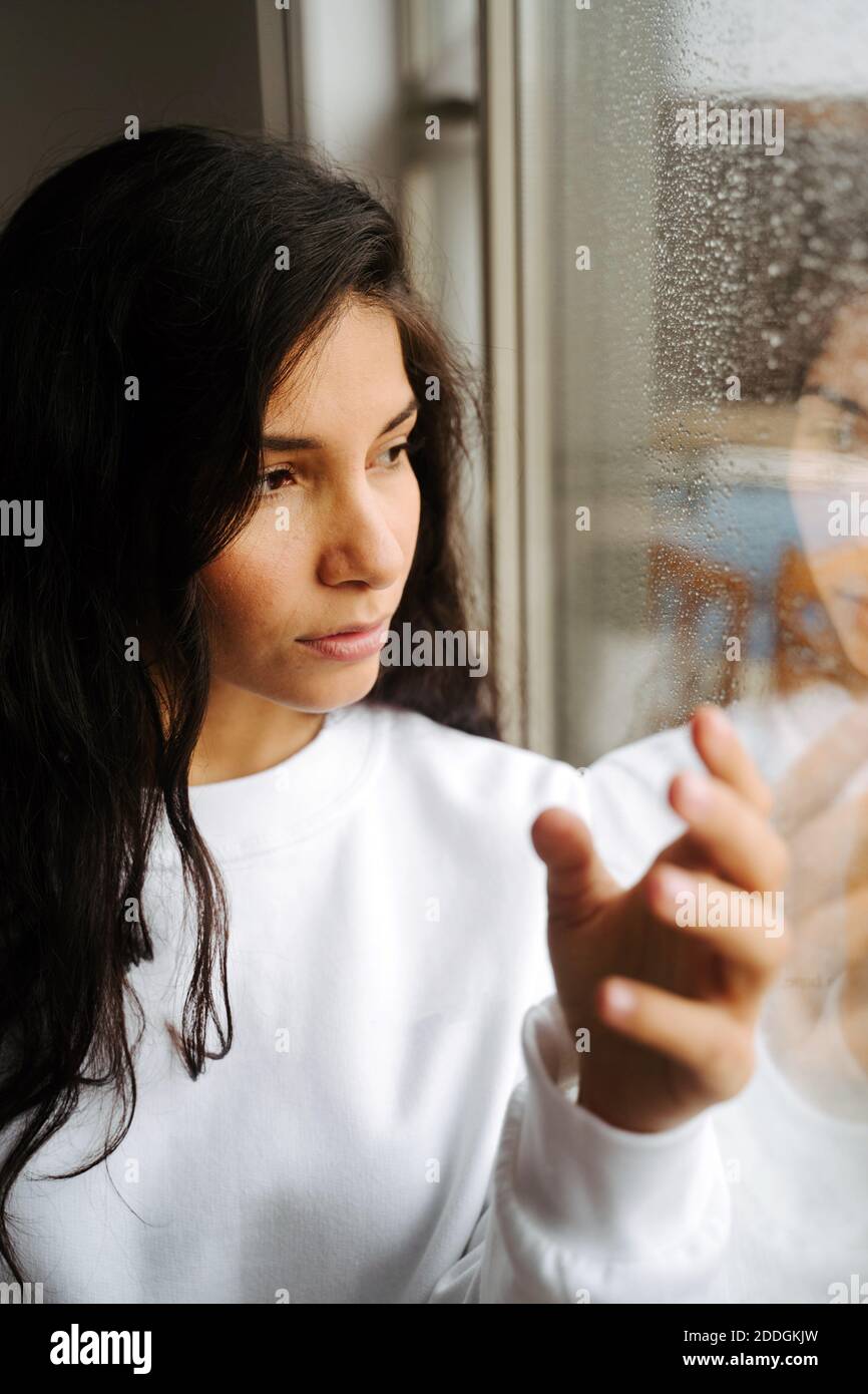 Young thoughtful ethnic female gently touching window with raindrops ...