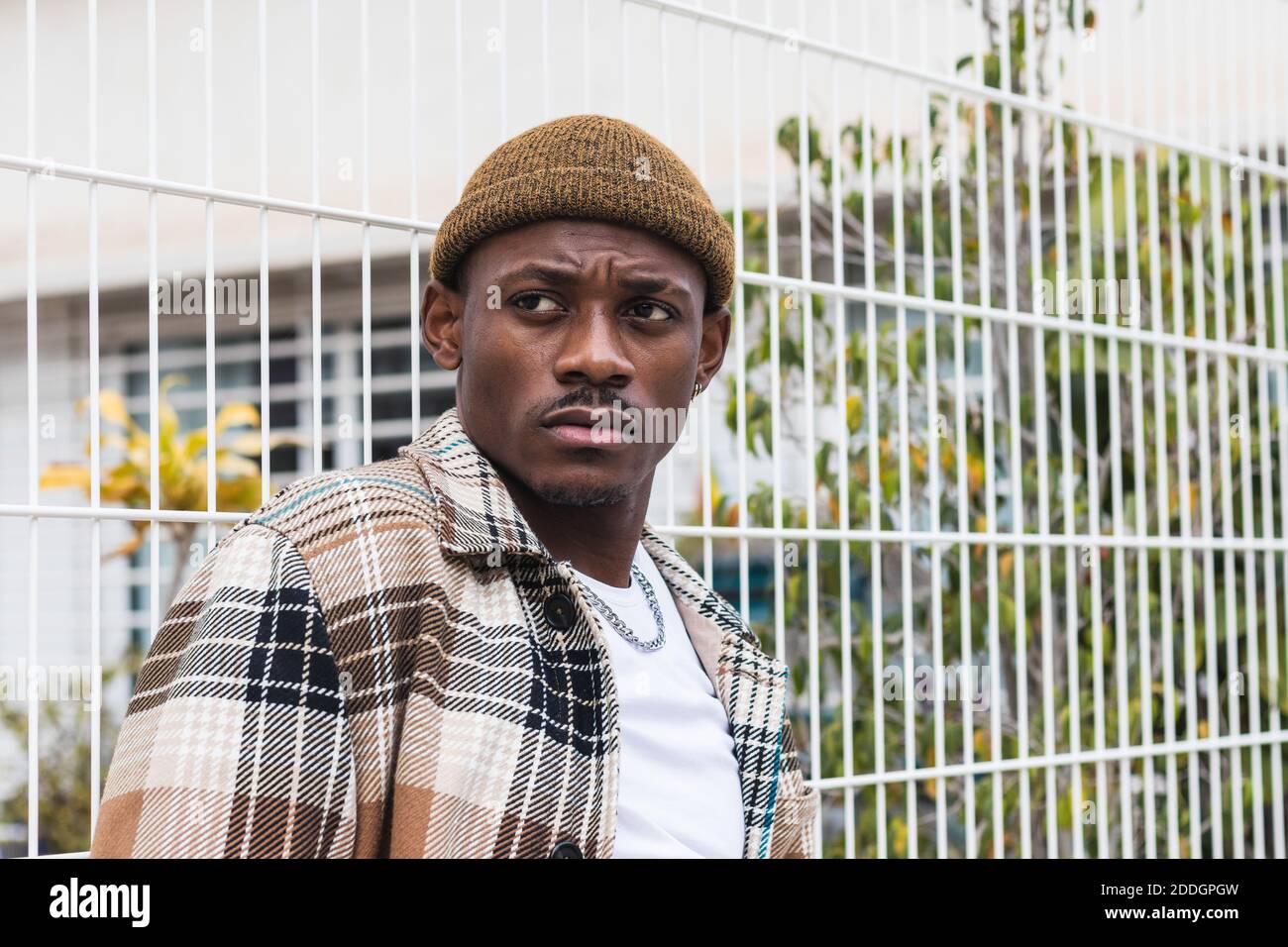 Young black man in stylish checkered shirt and hat leaning on net fence ...