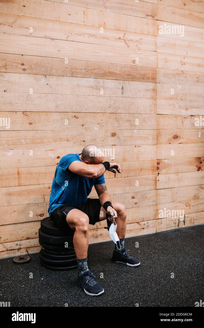 Male athlete with mask sitting on barbell weights while having break ...