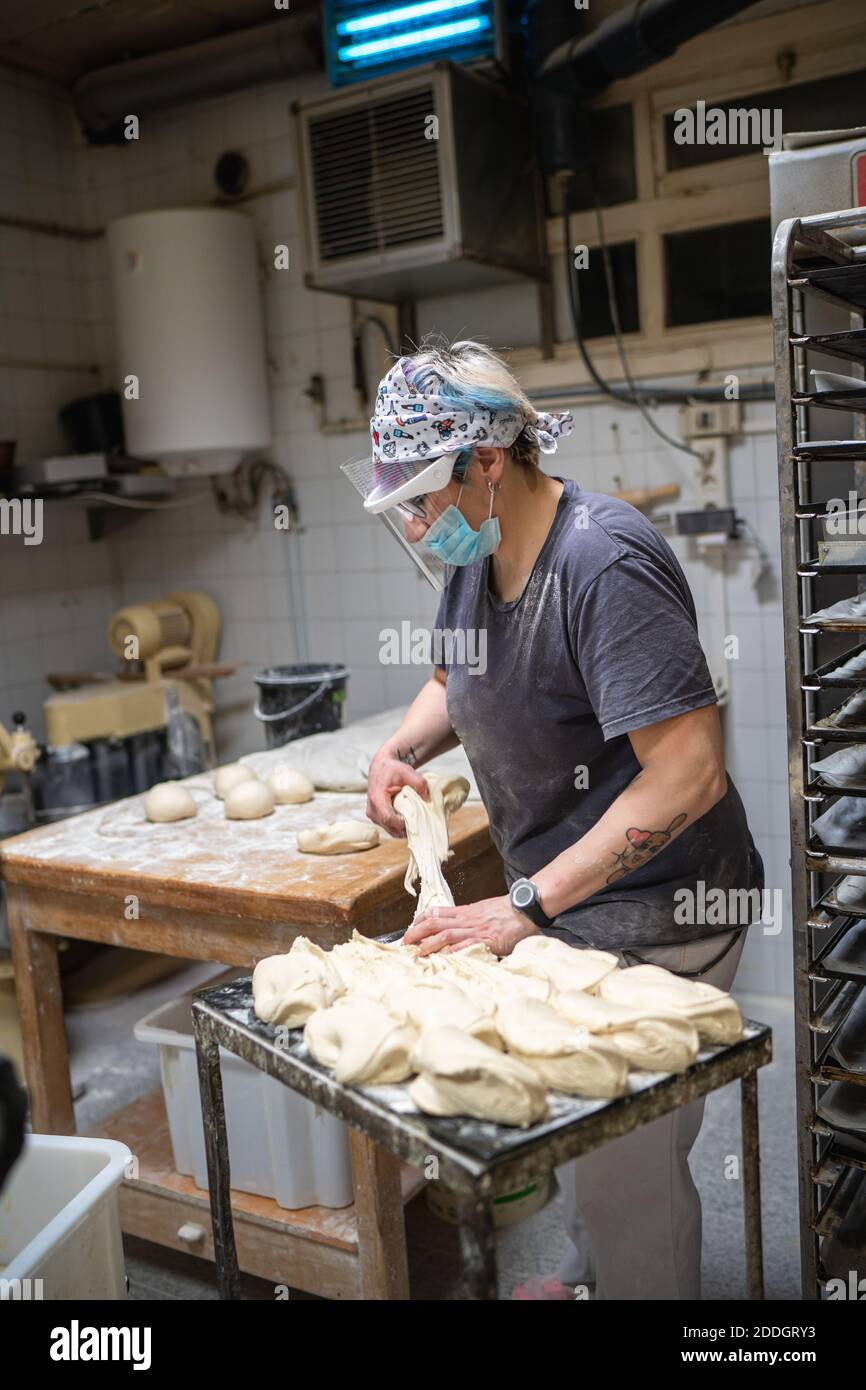 Side view of female baker in mask and shield standing at table in ...