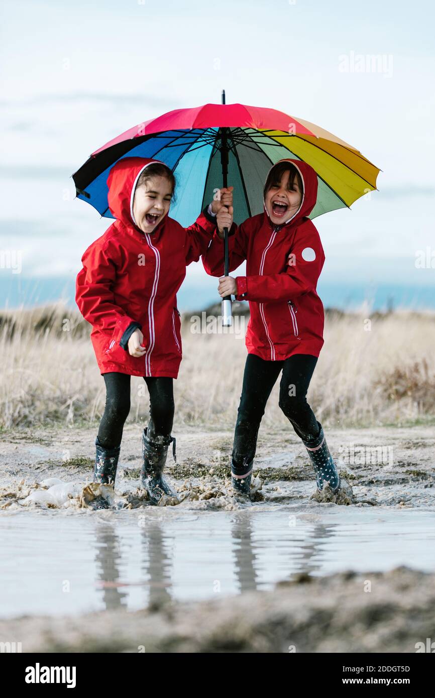 Excited sisters with colorful umbrella and in rubber boots playing in ...