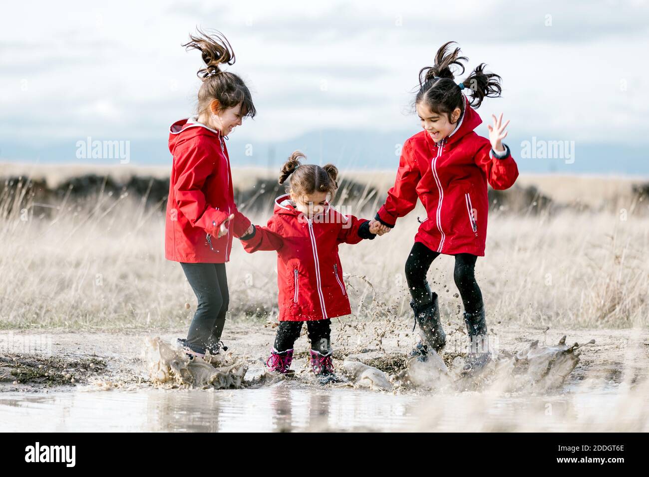 Delighted sisters holding hands and jumping in dirty puddle while ...