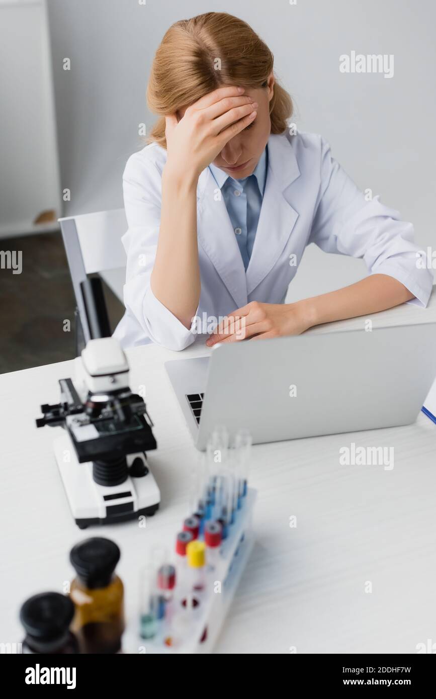 sad scientist in white coat covering face near laptop and microscope on ...