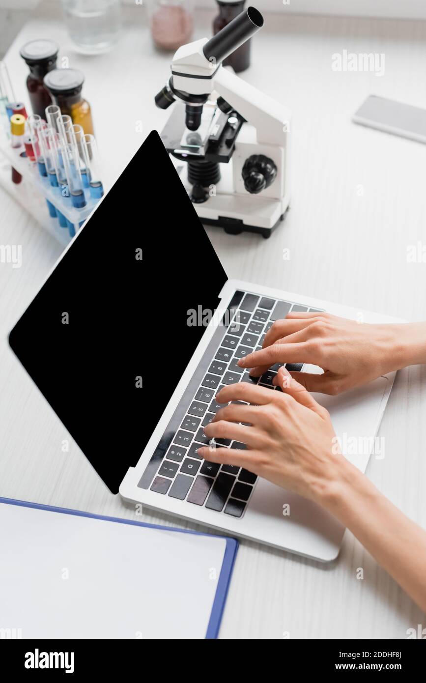 partial view of scientist typing on laptop with blank screen near ...
