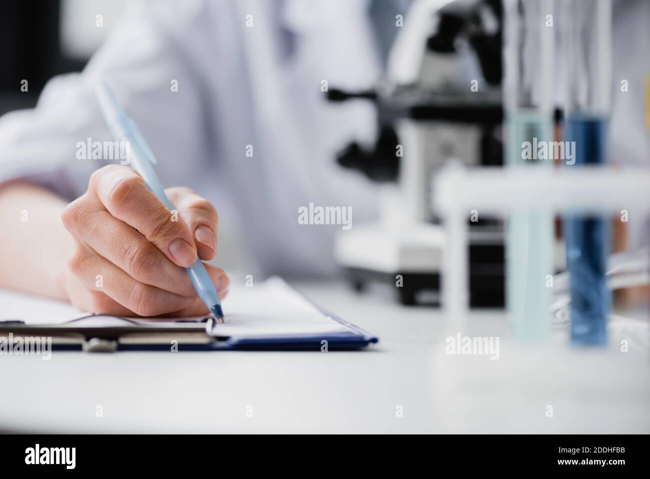 partial view of nurse writing on clipboard near microscope on blurred ...