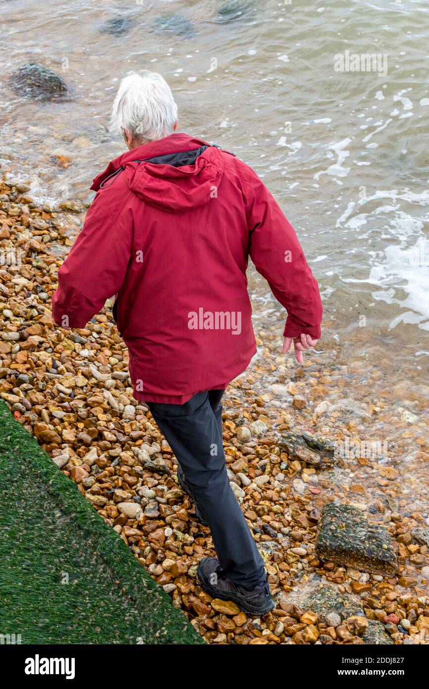 older grey haired man walking along a beach on the shingle with the sea ...