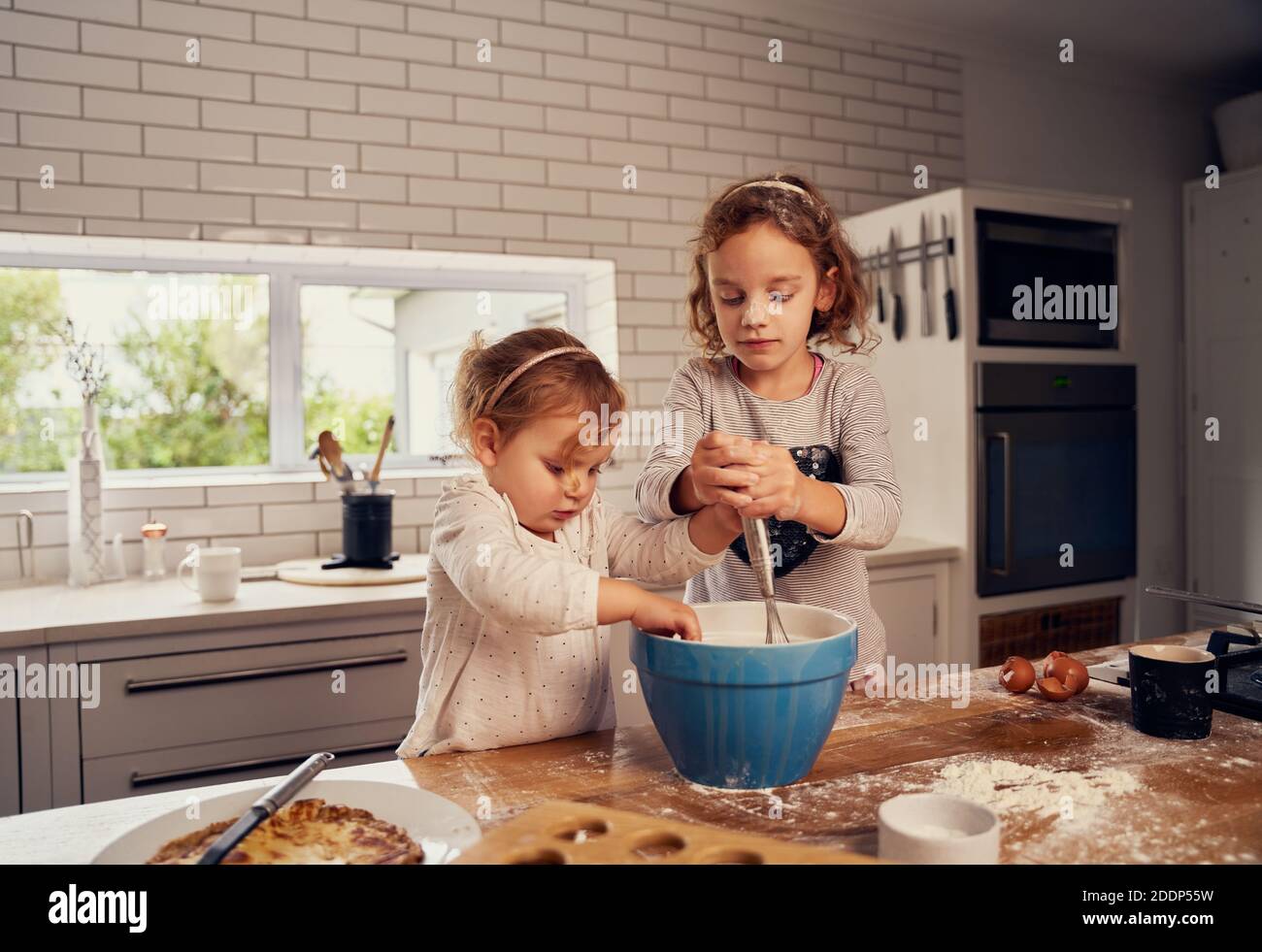 Little sisters enjoying making dough with eggs for baking in modern ...