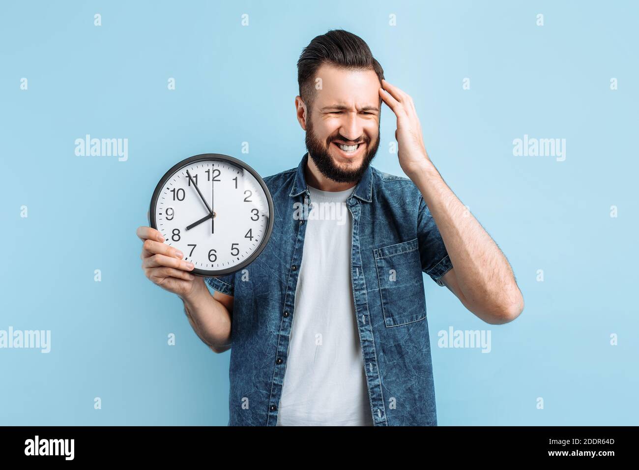 An exhausted, late- arriving young man with a beard holds a wall clock ...
