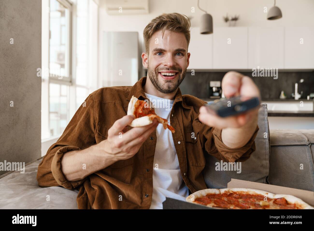 Handsome cheerful guy eating pizza while watching tv and using remote ...