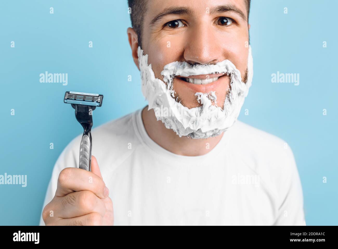 A handsome young man with a beard stands on a blue background with shaving foam on his face ...