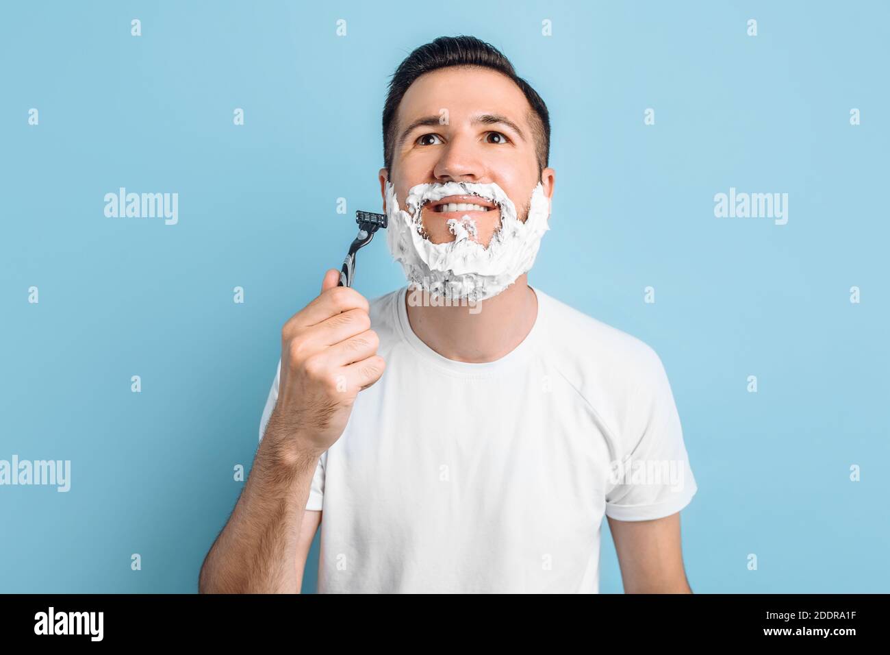 A handsome young man with a beard stands on a blue background with shaving foam on his face ...