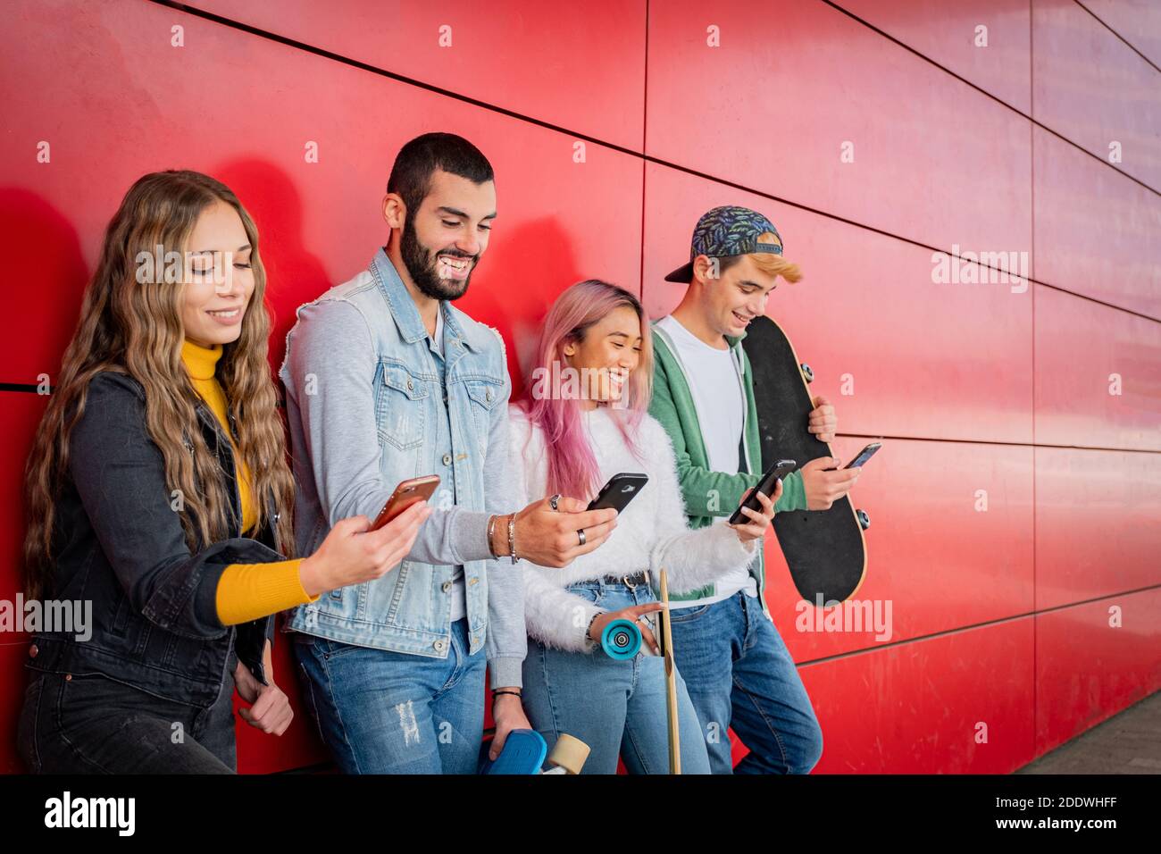 Group of young friends stare at their cell phones while standing against a wall in the city ...