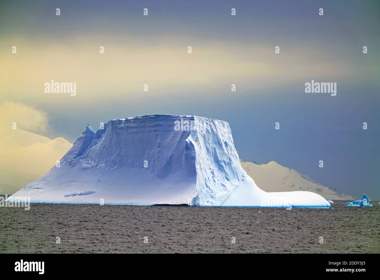 There is a light blue iceberg on the sea. Behind it there is a lump of ...