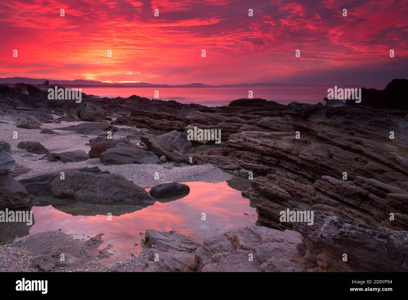 A blazing red sunset over a beach, with the clouds reflected in rock ...