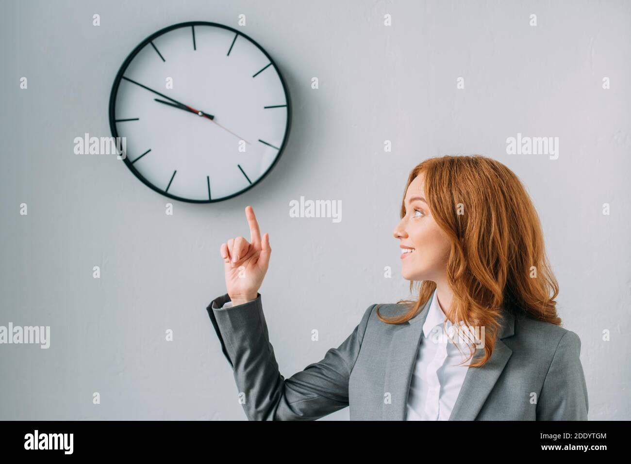 Smiling redhead businesswoman pointing with finger and looking at wall ...
