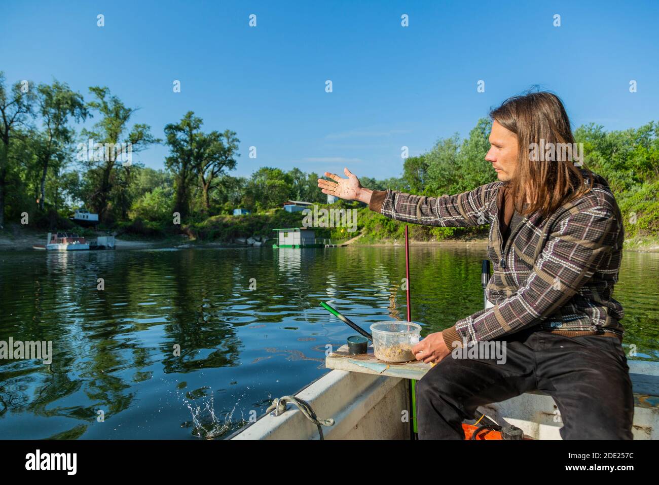 Fisherman throwing chum in lake. He is in white boat with trees around ...