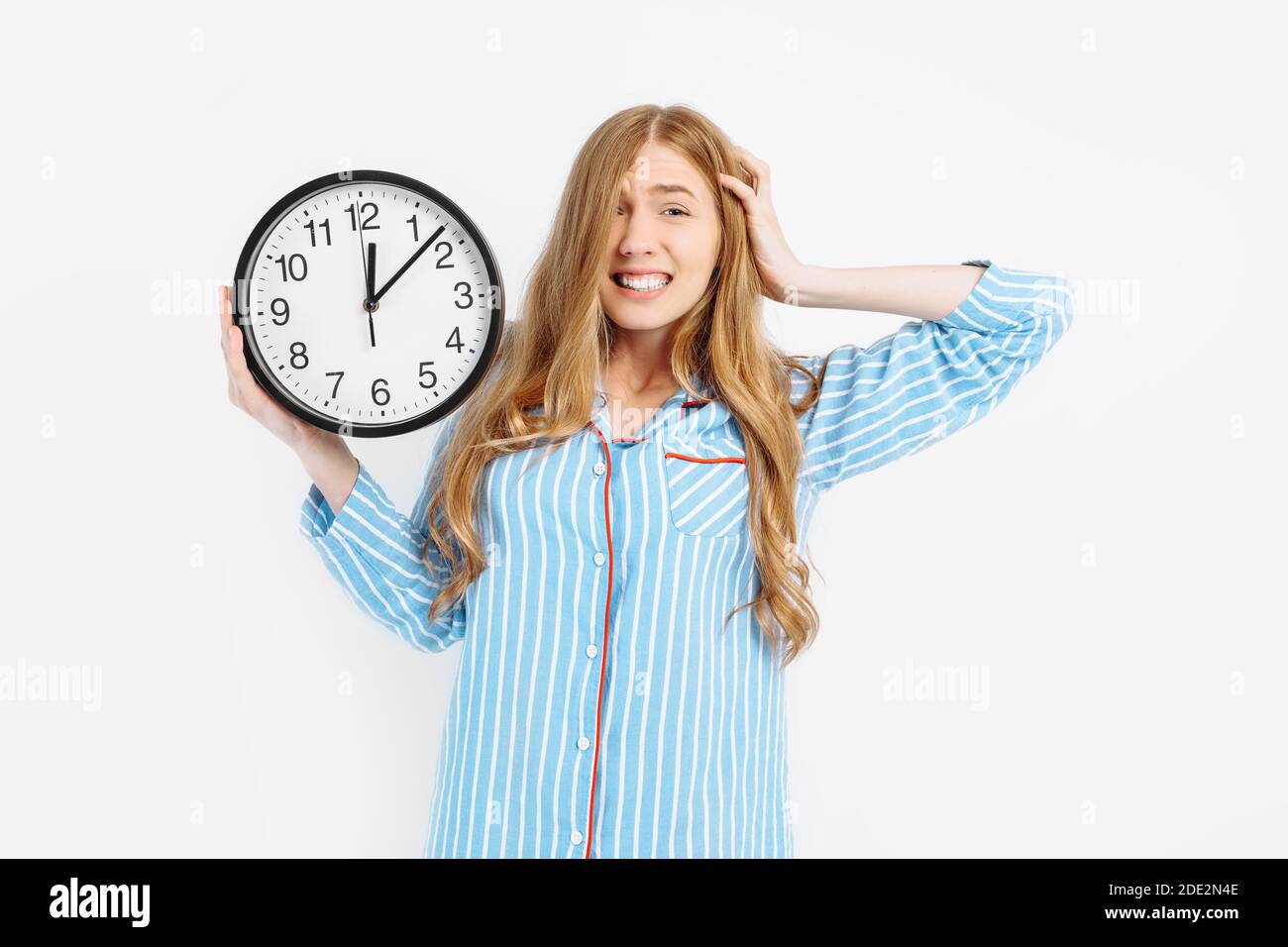 Portrait of young shocked confused girl in pajamas holding alarm clock ...