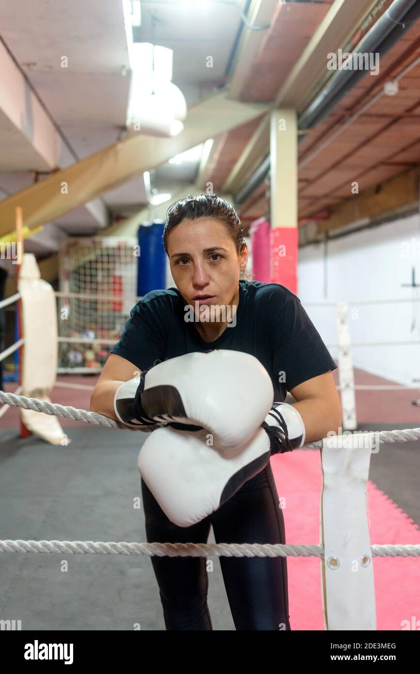 Real female boxer in gloves leaning on rope at ring before training ...