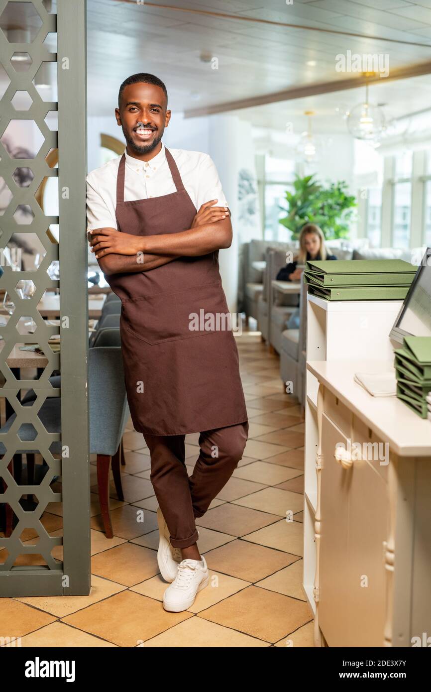 Cheerful young African waiter in workwear leaning against armchair by ...