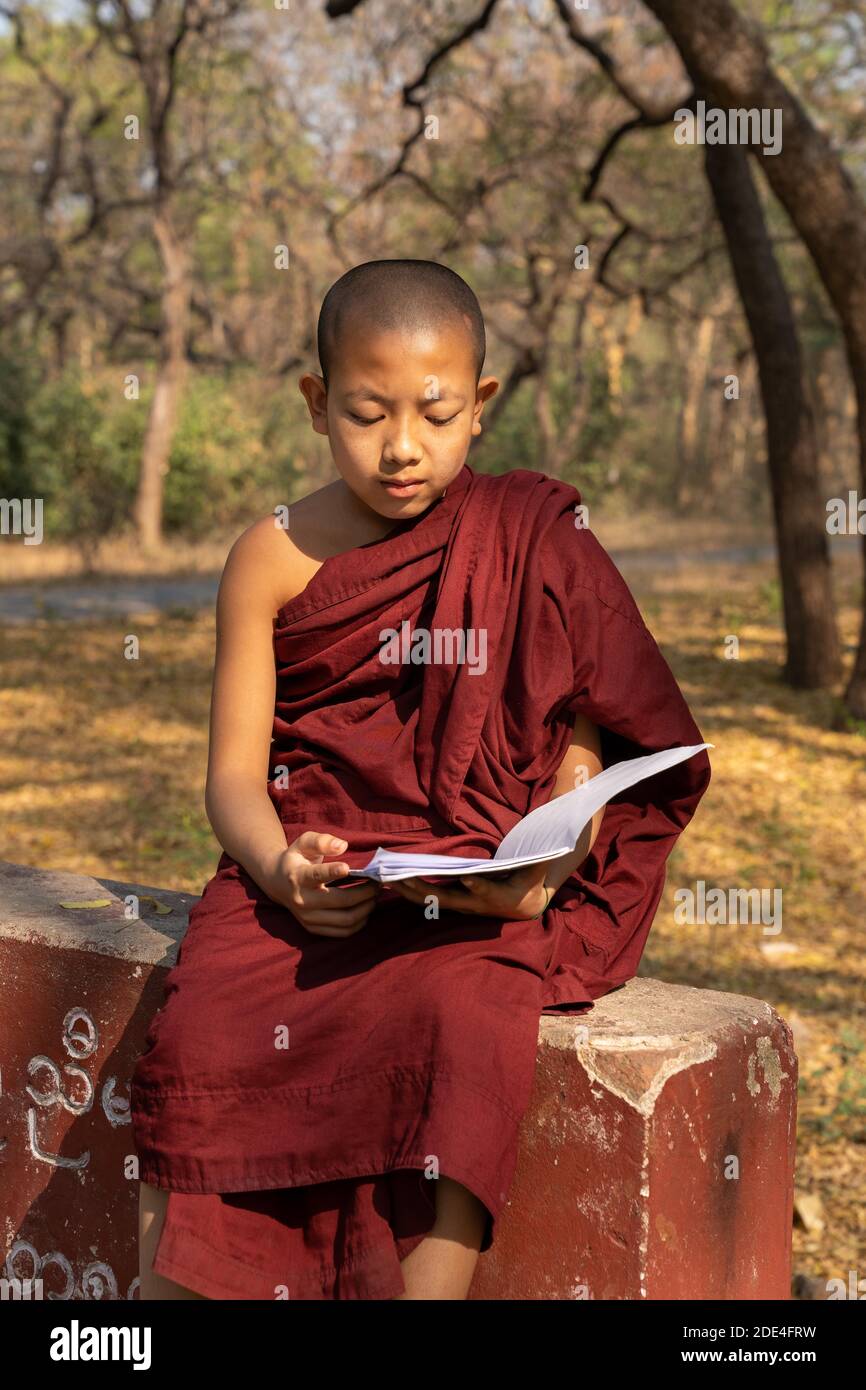 Child monk with red robe reading on a red wall, Shwe Hin Thar Monastery ...