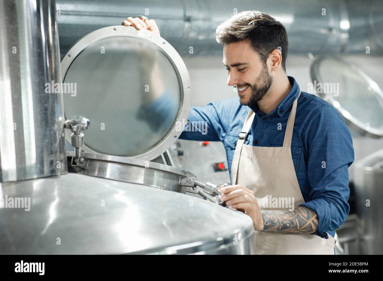 Smiling male worker or owner in apron opens lid of large boiler for ...