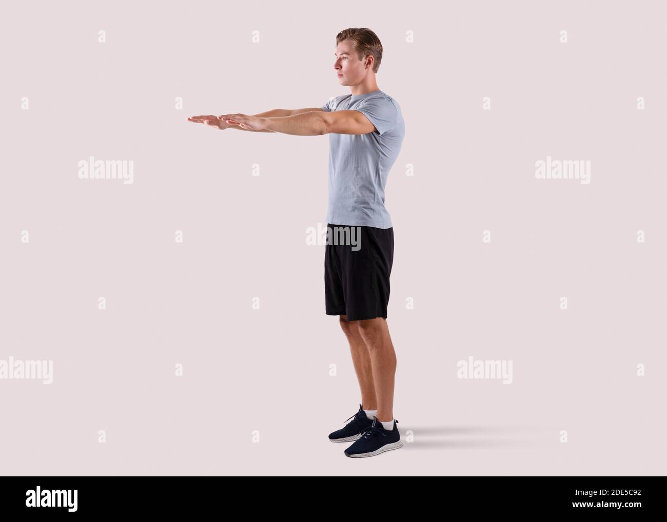 Strong millennial guy doing squats exercises on light studio background ...