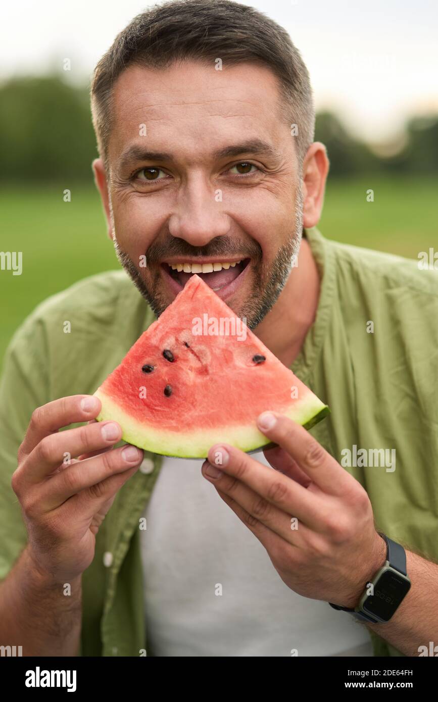 Happy young man looking at camera while eating watermelon in the green ...