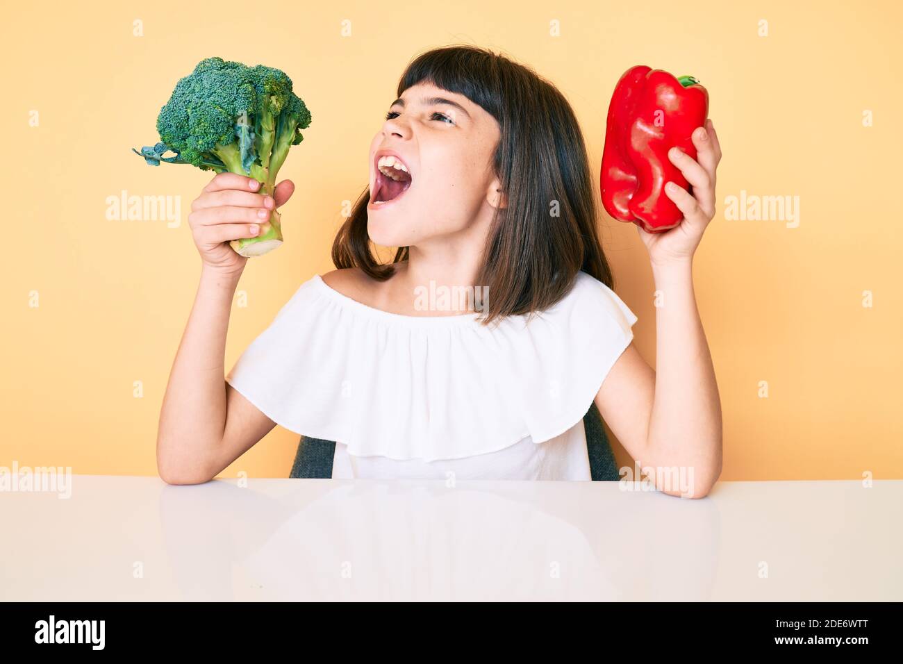 Young little girl with bang holding broccoli and red pepper angry and ...
