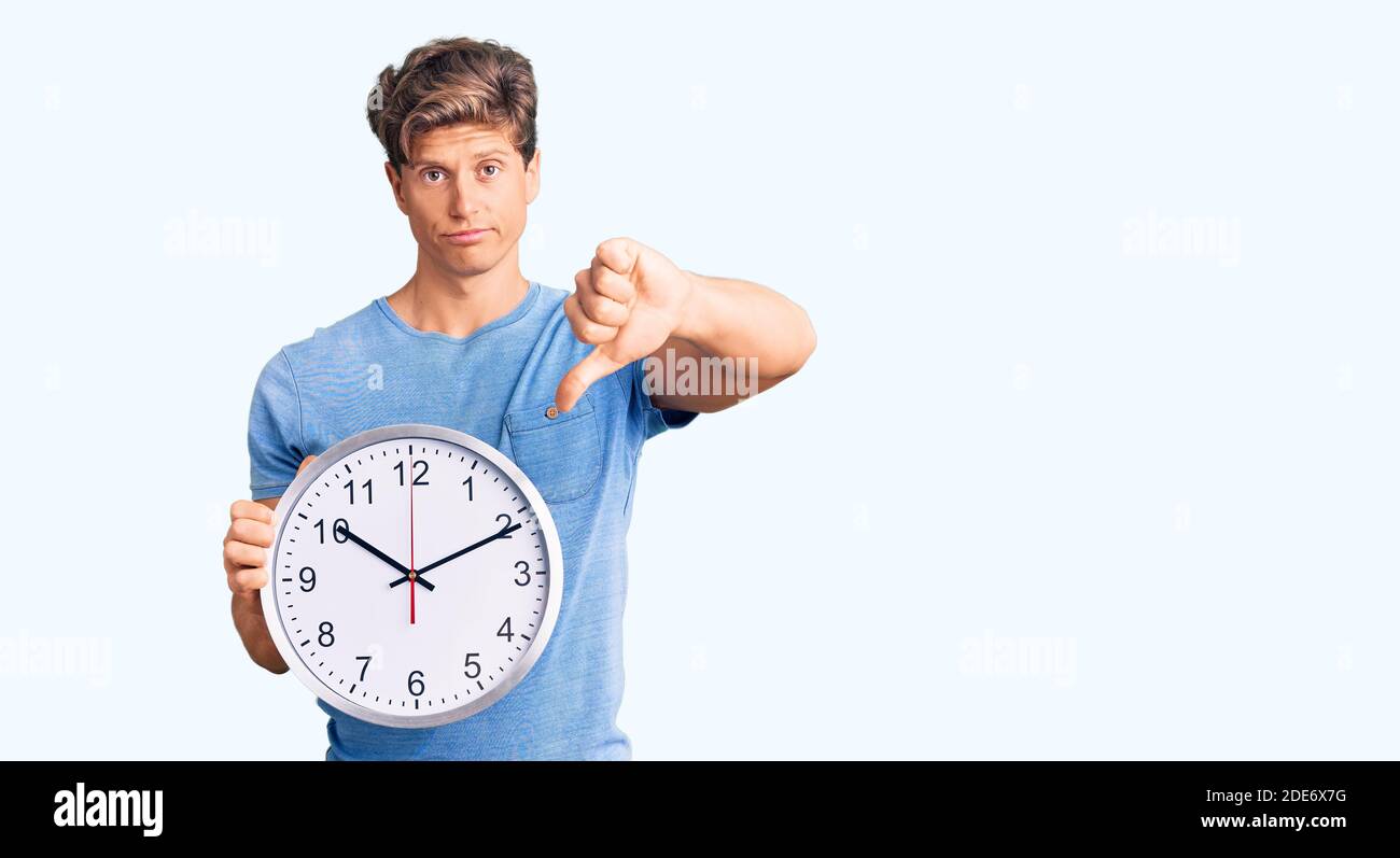 Young handsome man holding big clock with angry face, negative sign ...