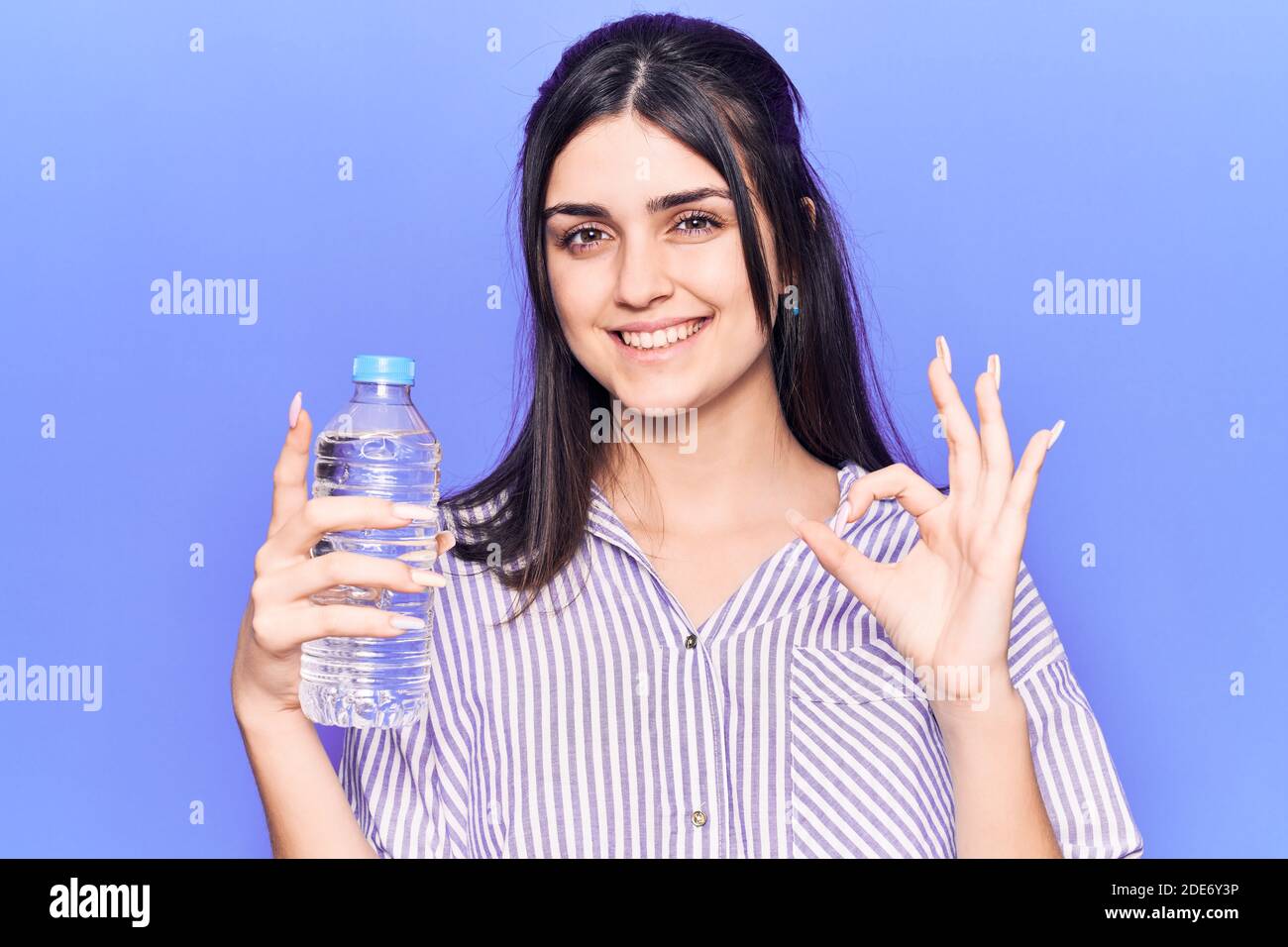 Young beautiful girl holding bottle of water doing ok sign with fingers ...