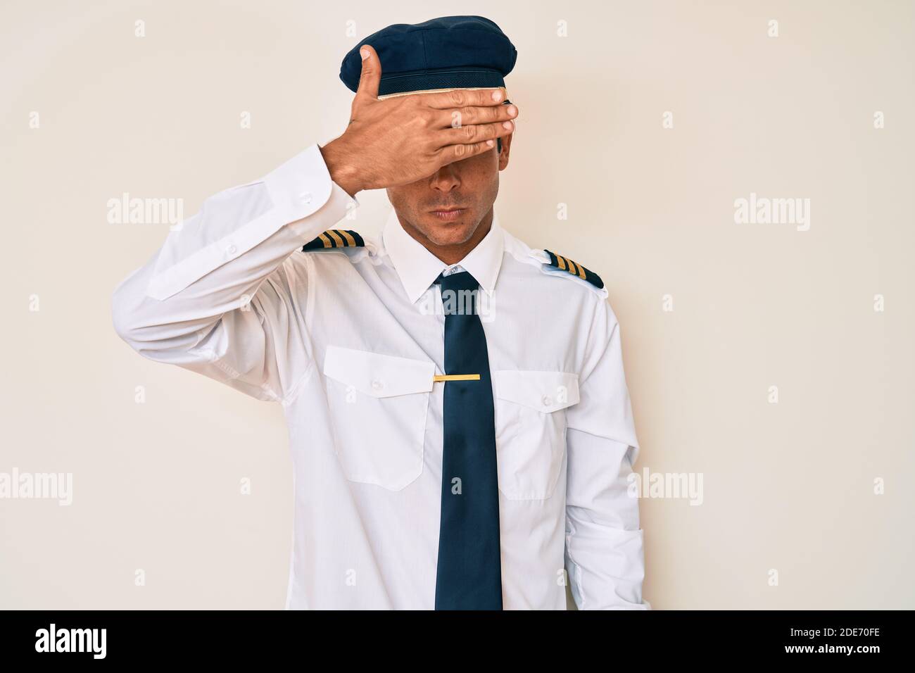 Young hispanic man wearing airplane pilot uniform covering eyes with ...