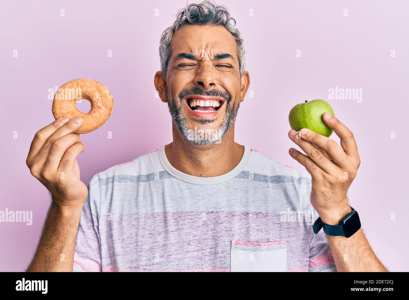 Middle age grey-haired man holding green apple and donut smiling and ...