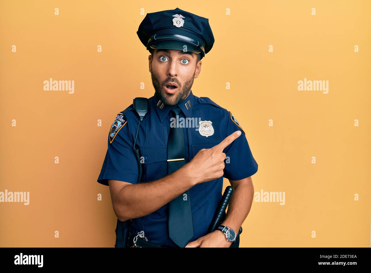Handsome hispanic man wearing police uniform surprised pointing with ...