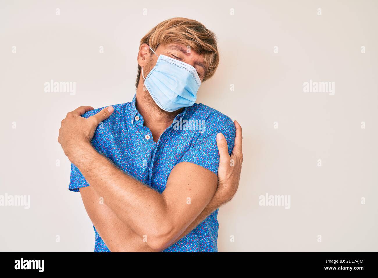 Young blond man wearing medical mask hugging oneself happy and positive ...