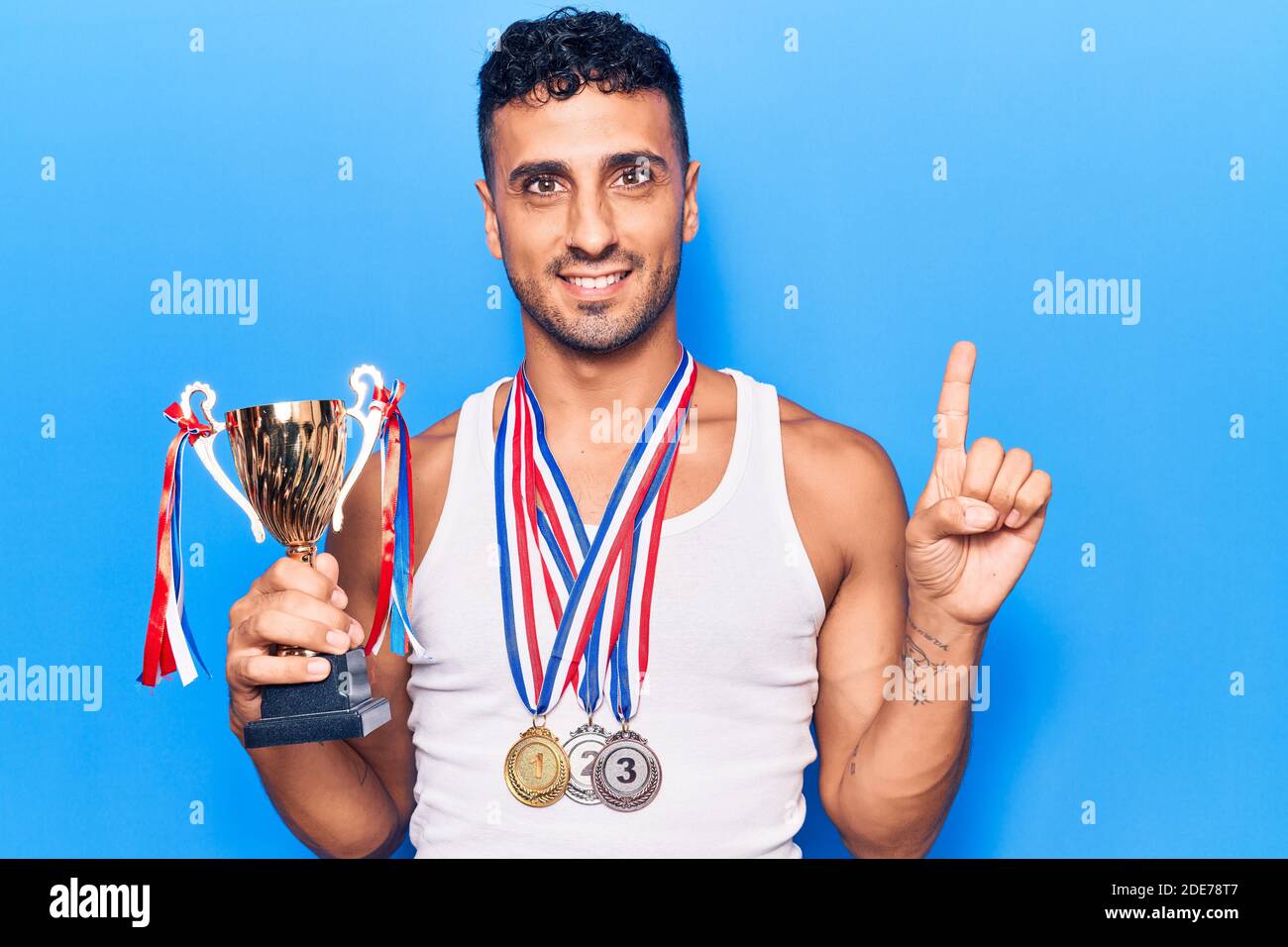 Young hispanic man wearing winner medals holding trophy smiling with an ...
