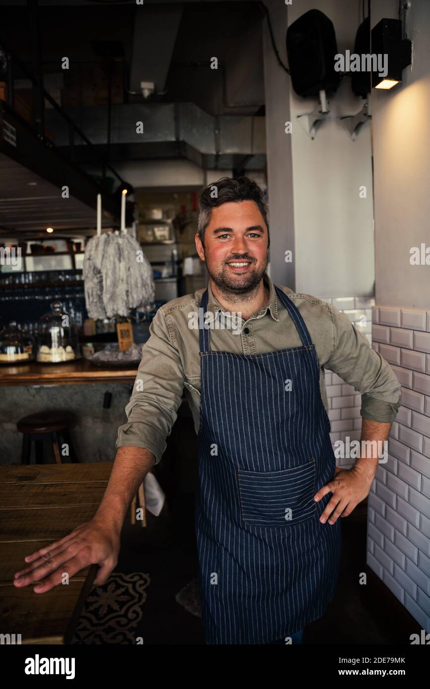 Smiling handsome caucasian waiter leaning on counter of funky cafe ...