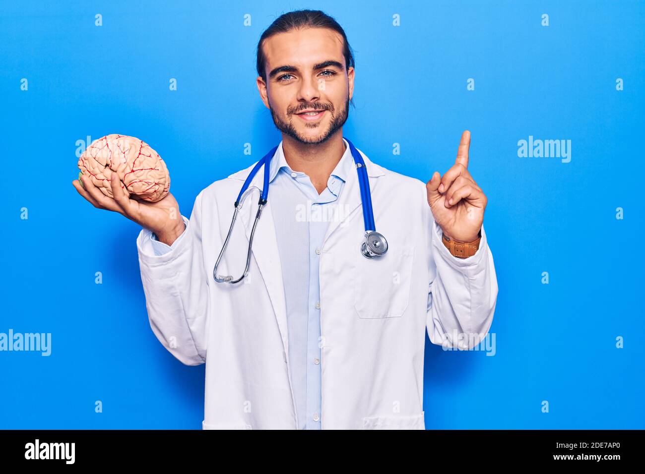 Young handsome man wearing doctor coat holding brain surprised with an idea or question pointing ...