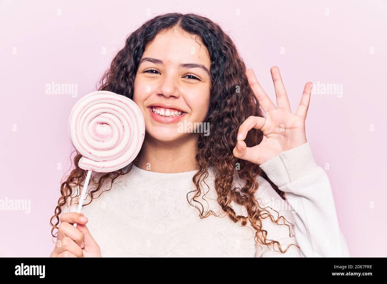 Beautiful kid girl with curly hair holding lollipop doing ok sign with ...