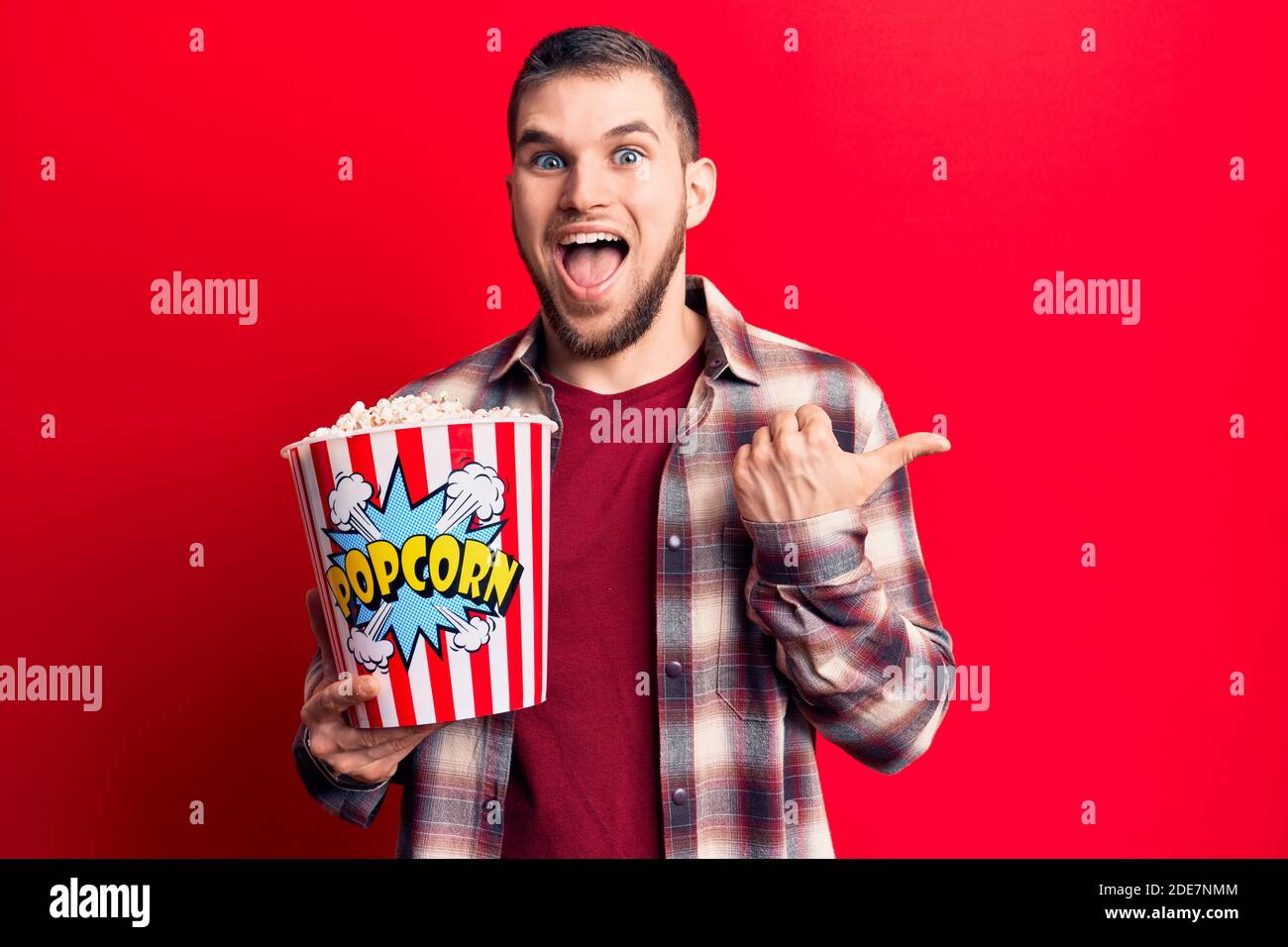 Young handsome man eating popcorn pointing thumb up to the side smiling ...