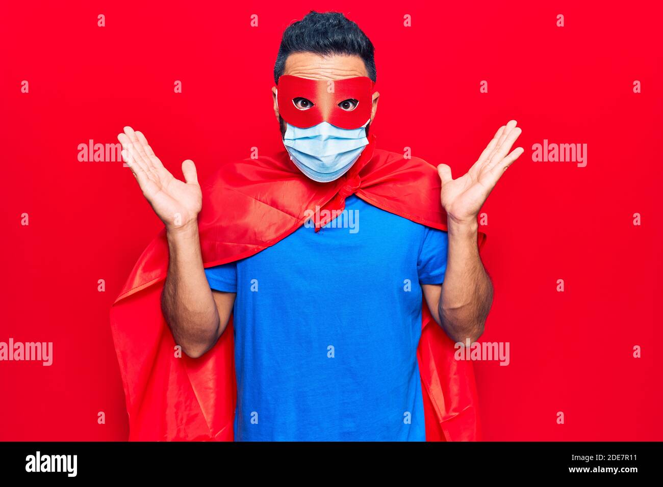 Young hispanic man wearing super hero costume and medical mask scared ...