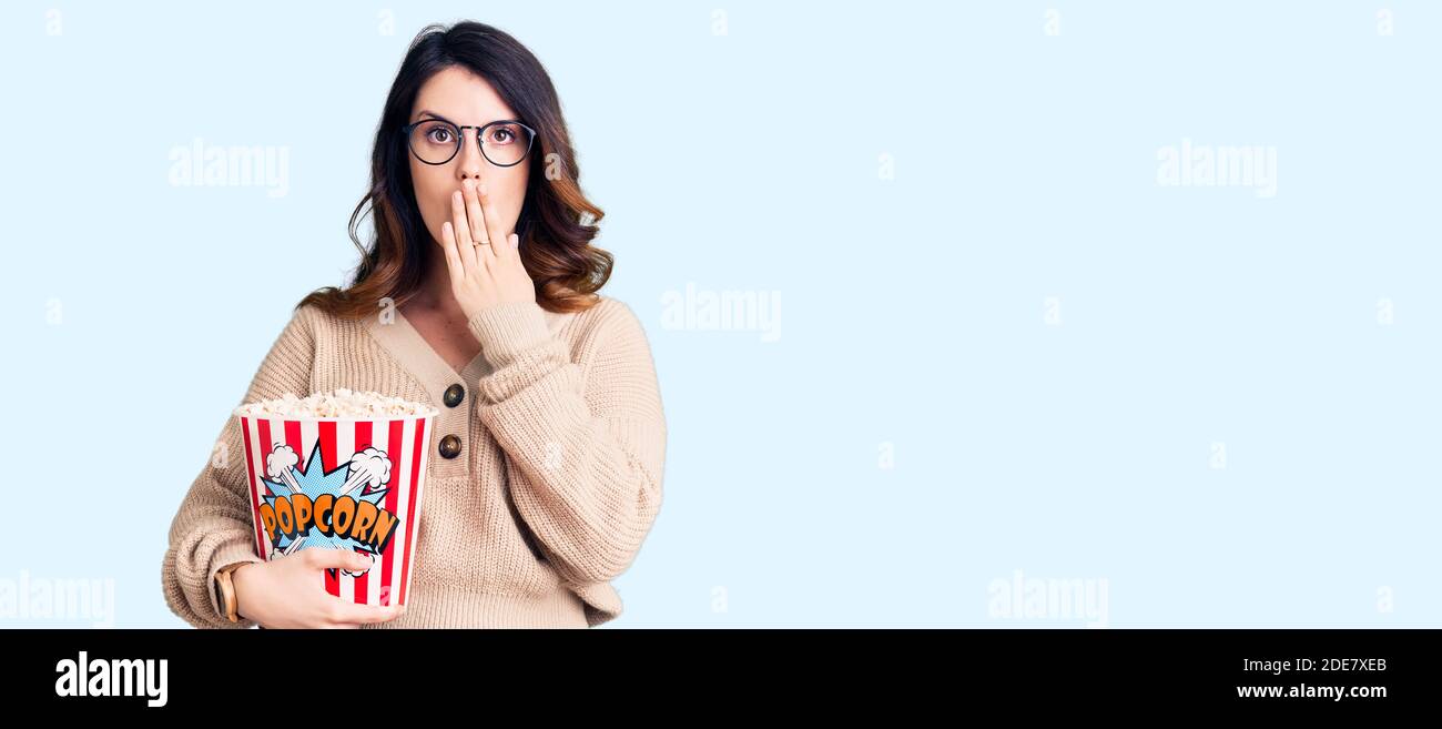 Beautiful young brunette woman eating popcorn covering mouth with hand ...