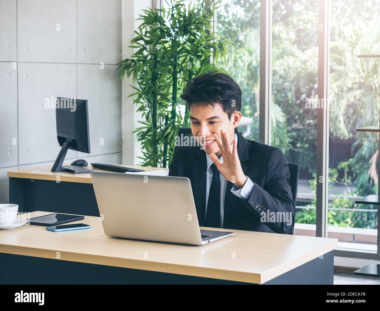 Young smiling Asian businessman in suit greeting someone, wave to ...