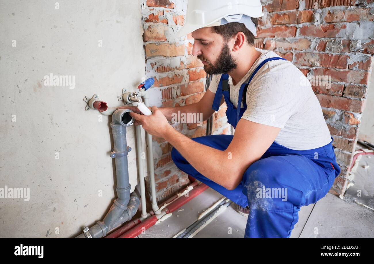 Young plumber, wearing blue uniform and white helmet working with ...